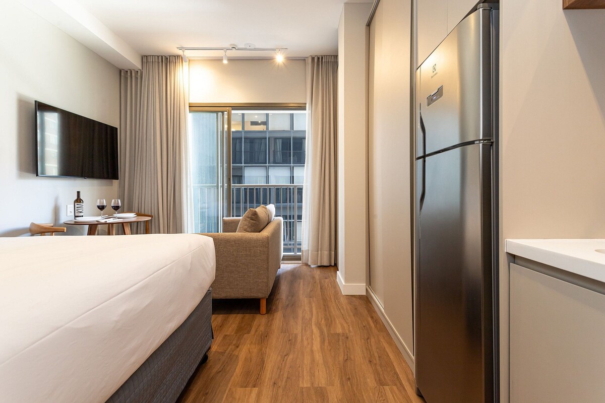 Quarto de hotel com uma cama, televisão, mesa de jantar com dois copos, uma cadeira, sofá, frigorífico e janela com cortinas.