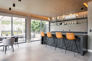 Uma área de bar moderna com pé-direito alto, janelas amplas e vista para edifícios e vegetação.