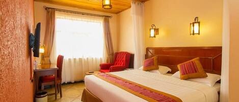 Quarto de hotel com teto de madeira, uma cama grande com colcha vermelha e branca, duas luminárias fixadas na parede, uma cadeira vermelha, uma mesa pequena com abajur e uma janela com cortinas.