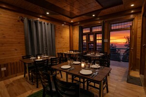 Um restaurante com paredes e teto de madeira, mesas preparadas para refeições e vista para o pôr do sol através de grandes janelas.