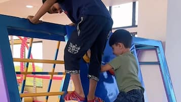 Uma criança escala uma estrutura de escalada azul com corrimãos amarelos numa área de recreação coberta.