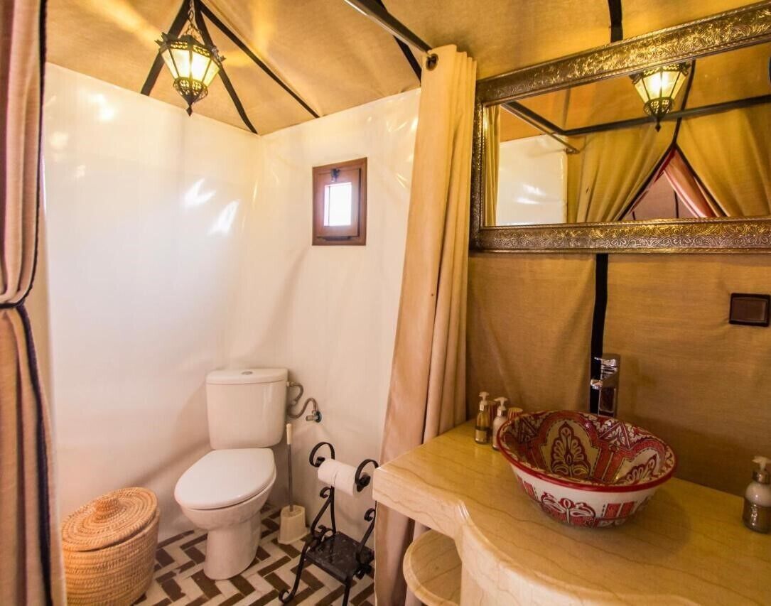 Casa de banho compacta com sanita branca, toucador de madeira, lavatório em taça decorativo, rolo de papel higiénico suspenso e uma pequena janela.