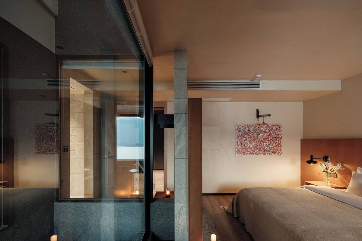 Quarto moderno com divisória de vidro, uma cama, mesa de cabeceira, obra de arte na parede e ar-condicionado instalado no teto.