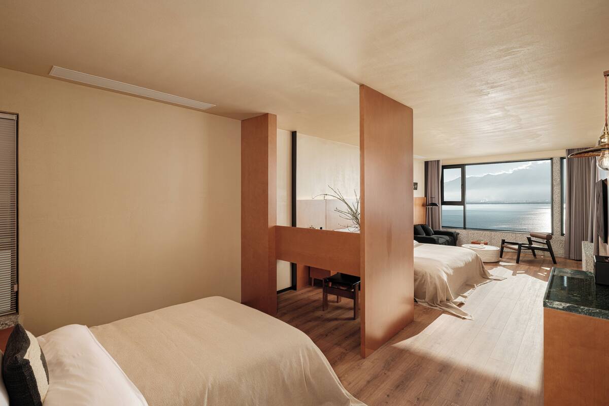 Quarto de hotel com duas camas, uma escrivaninha, uma cadeira e uma janela grande com vista para o mar.