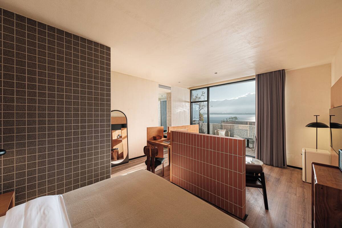 Quarto de hotel com uma cama grande, uma escrivaninha, uma cadeira, uma mesinha, um espelho, uma janela com vista e um painel divisório.