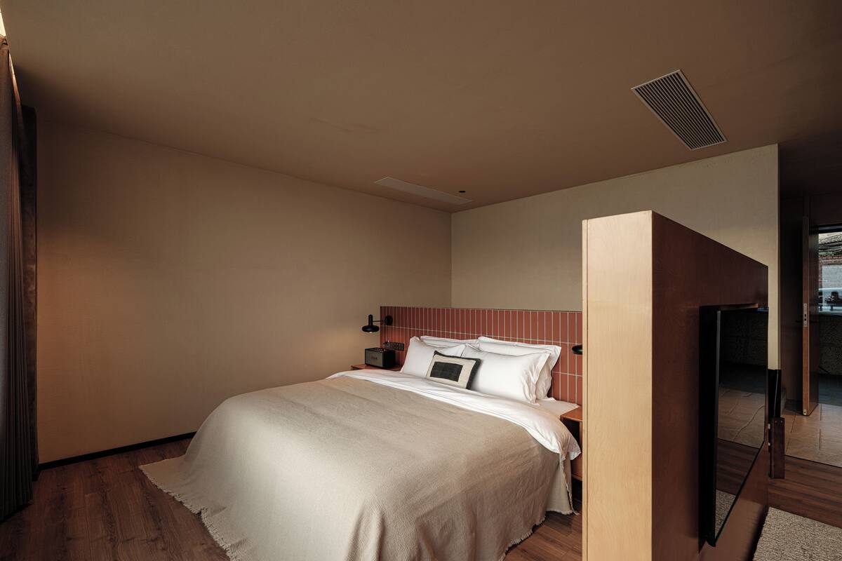 Quarto de hotel com cama, mesa de cabeceira, abajur e piso de madeira.