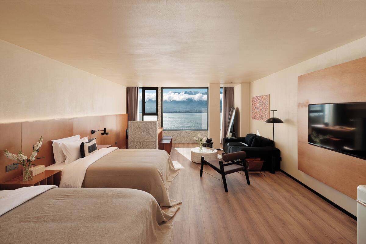 Quarto de hotel com duas camas, uma escrivaninha, uma cadeira, televisão e uma janela grande com vista para montanhas e água.