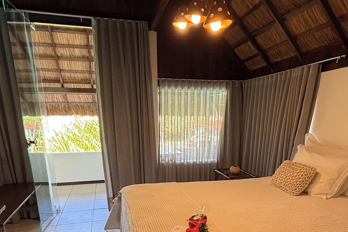 Quarto de hotel com uma cama grande, porta de vidro que dá para área externa e teto de palha.