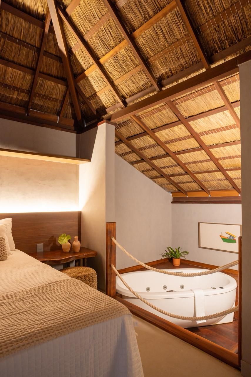 Quarto com telhado de palha, uma cama, uma mesinha com uma planta e uma banheira branca.
