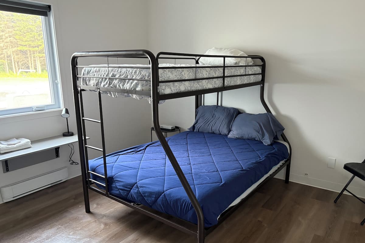 Uma cama de solteiro com colchão e almofadas azuis, uma mesinha de cabeceira, uma janela com vista para árvores e um chão de madeira.