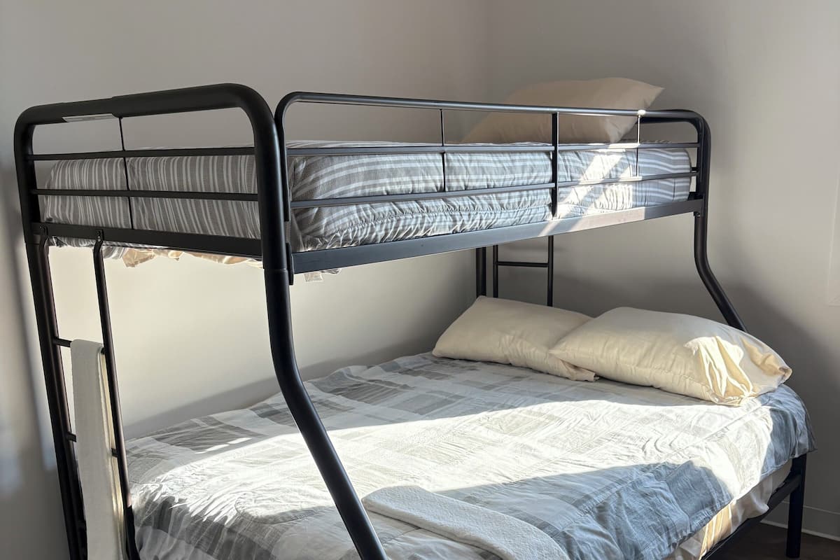 Uma cama beliche com colchão e travesseiros, piso de madeira e parede lisa.