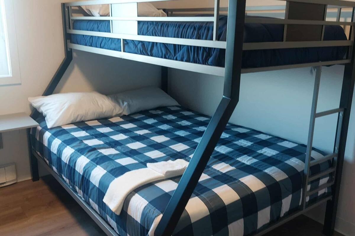 Uma cama beliche com colcha xadrez azul e branca, um travesseiro branco e uma janela à esquerda.