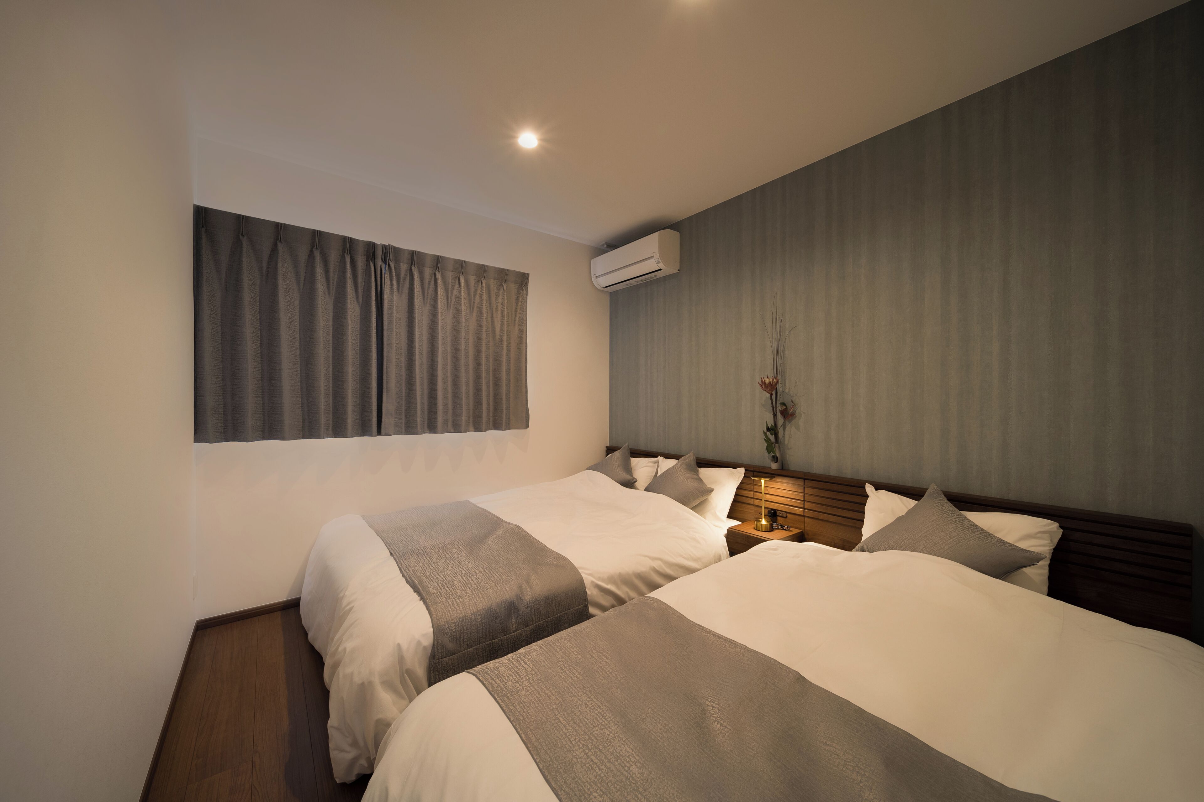 Quarto de hotel com duas camas, cabeceira de madeira, ar condicionado fixado na parede e mesa de cabeceira com luminária.