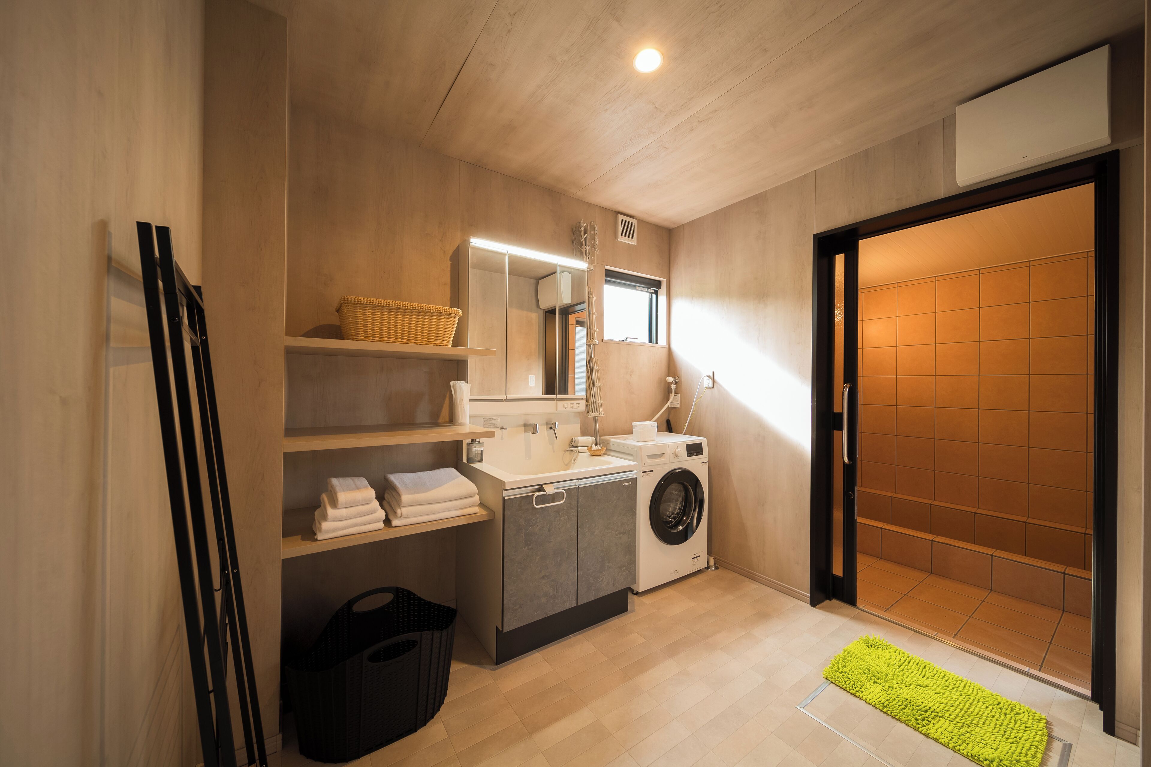 Casa de banho compacta e moderna com máquina de lavar roupa, uma pia e um espelho. Há uma prateleira com toalhas e uma cesta. Um tapete verde está no chão.