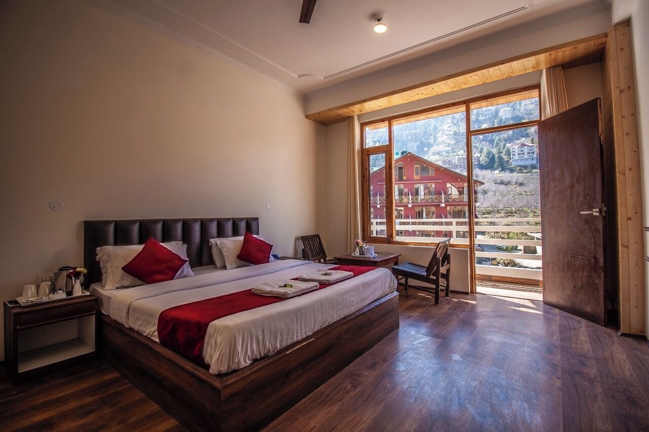 Uma cama grande com cabeceira de madeira, uma mesa de cabeceira e vista para um edifício vermelho através de uma janela ampla.