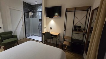 Quarto de hotel com cama, secretária, cadeira, televisão, chuveiro e guarda-roupa.