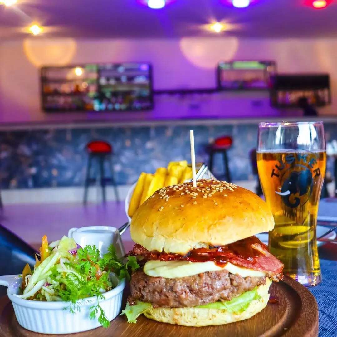 Um hambúrguer com pão de gergelim, bacon, alface e uma porção de salada de repolho, acompanhado de uma cerveja e batatas fritas.