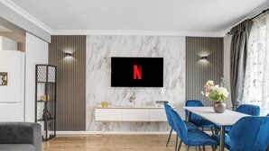 Uma sala de estar moderna com televisão de tela plana fixada em uma parede de mármore, uma área de jantar com cadeiras azuis e um sofá cinza.