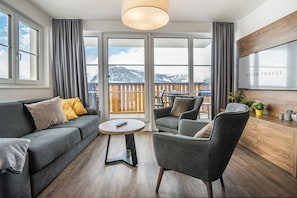 Quarto de hotel moderno com sofá, poltronas, uma mesa pequena e varanda com vista para montanhas cobertas de neve.