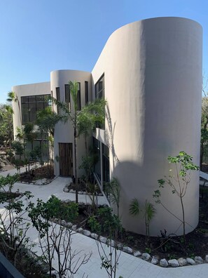 Um edifício moderno com fachada curva, janelas amplas e um pátio ajardinado com diversas plantas.