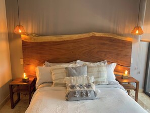 Quarto com cabeceira de madeira, várias almofadas, mesas de cabeceira com abajures e uma janela grande.