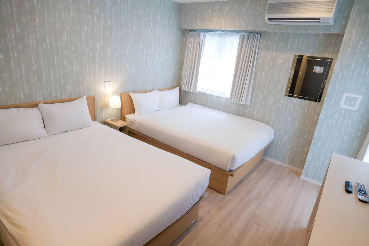 Quarto de hotel com duas camas, uma mesa de cabeceira com abajur, unidade de ar condicionado, janela com cortinas e um pequeno espelho na parede.