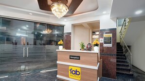 Uma área de recepção de hotel com balcão de recepção identificado como "Tab Hotels", um lustre, uma escadaria e uma placa indicando os horários de check-in e check-out.