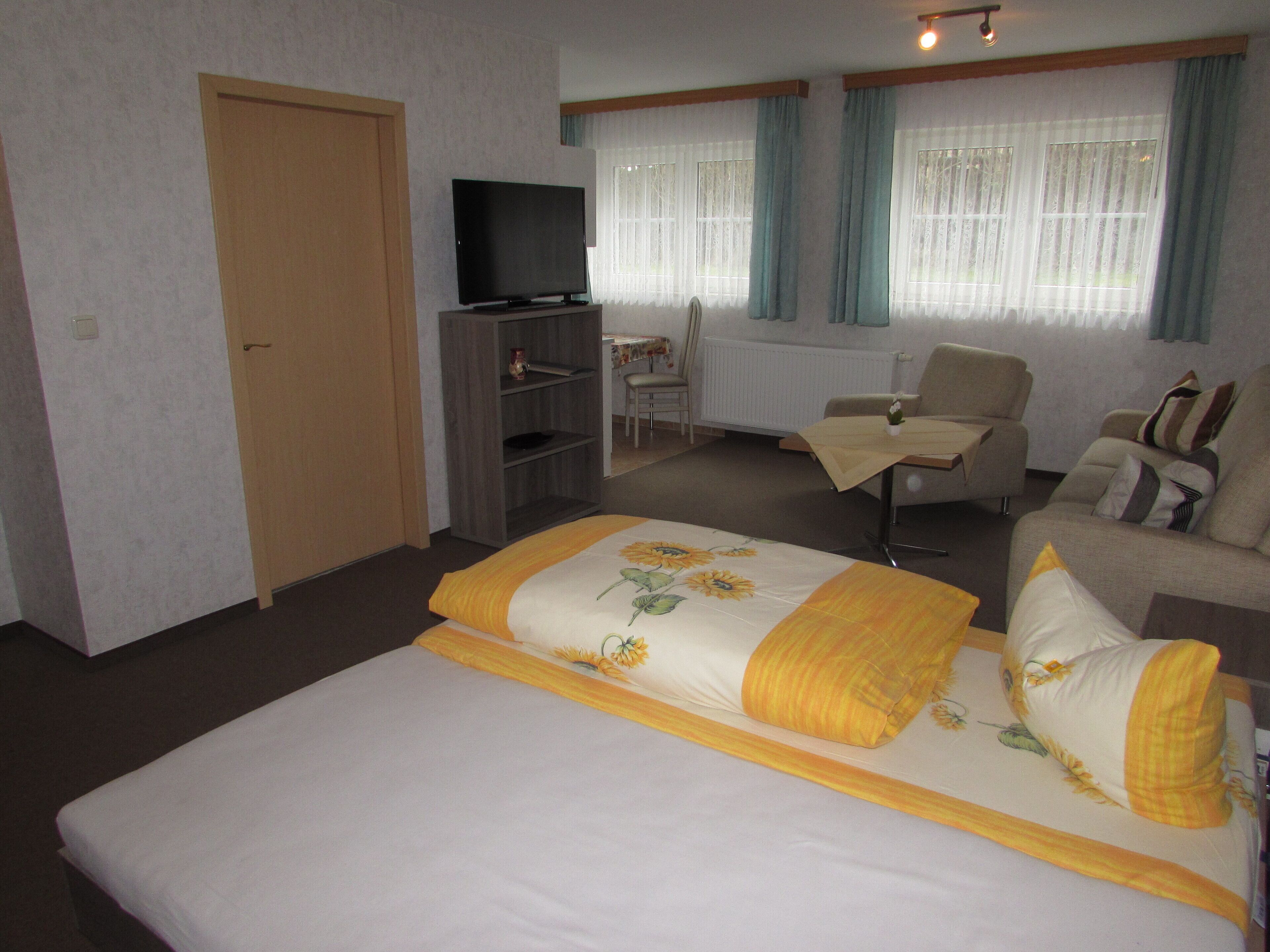Quarto de hotel com cama, televisão, poltrona, sofá e janela com cortinas.