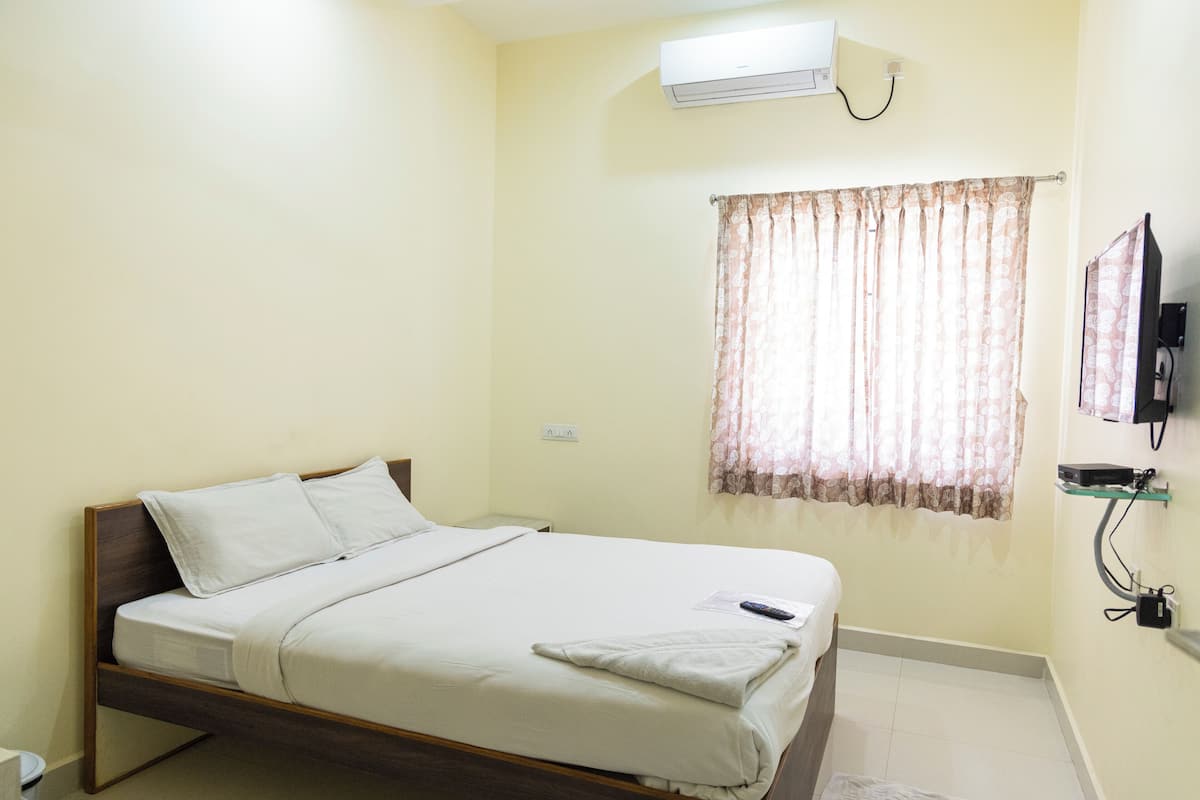 Cama bem arrumada com controle remoto, ar-condicionado fixado na parede e janela com cortinas estampadas.