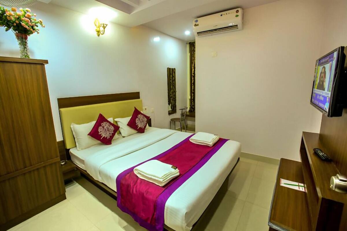 Quarto de hotel com uma cama, guarda-roupa de madeira, escrivaninha com cadeira, ar condicionado e televisão.