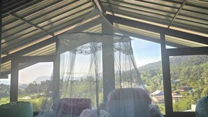 Quarto com cama coberta por mosquiteiro, janelas amplas e vista para uma paisagem verde.