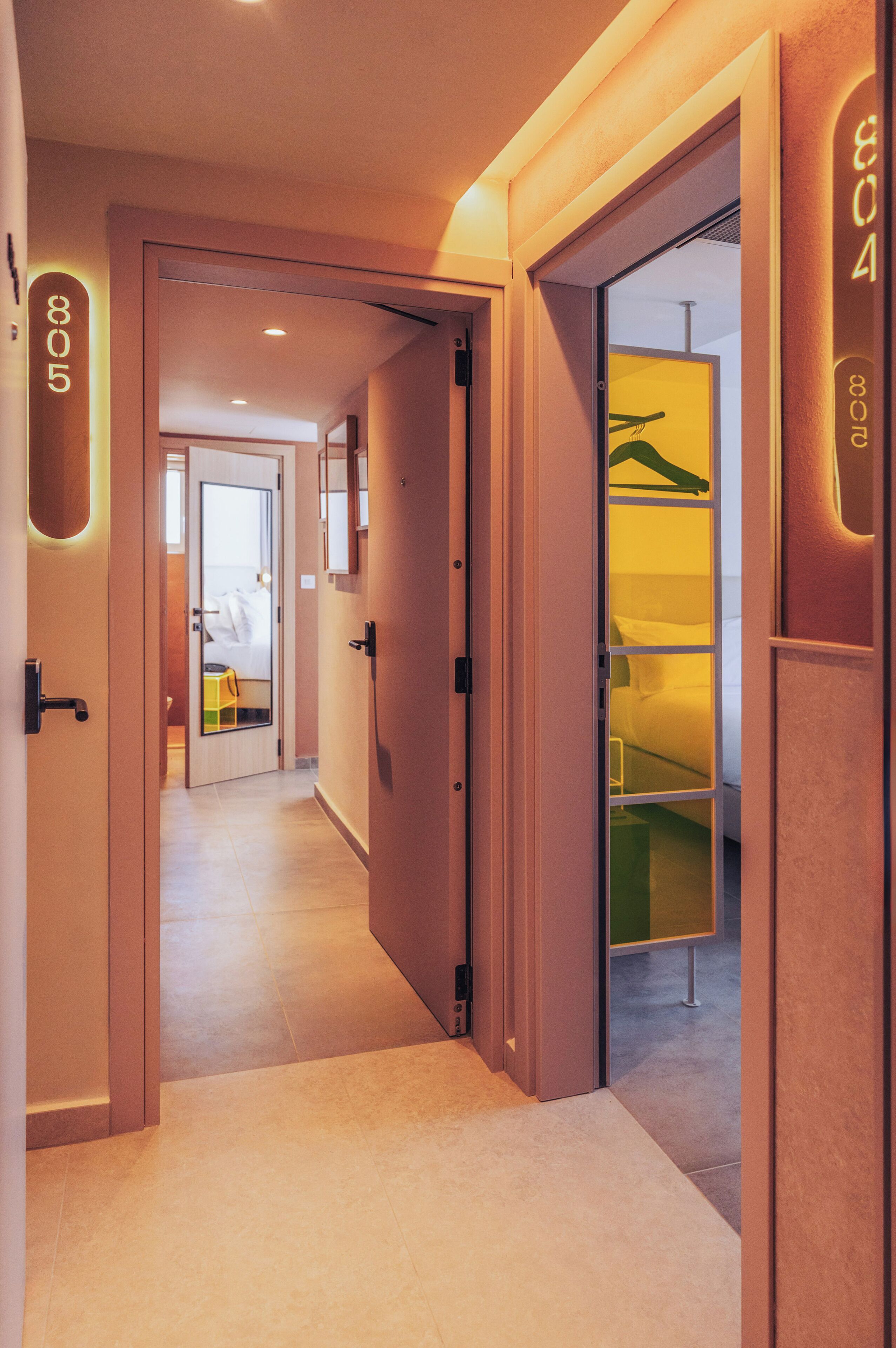 Corredor de hotel com design moderno, com uma porta com placa de número, um espelho e uma cama visível por uma porta aberta.