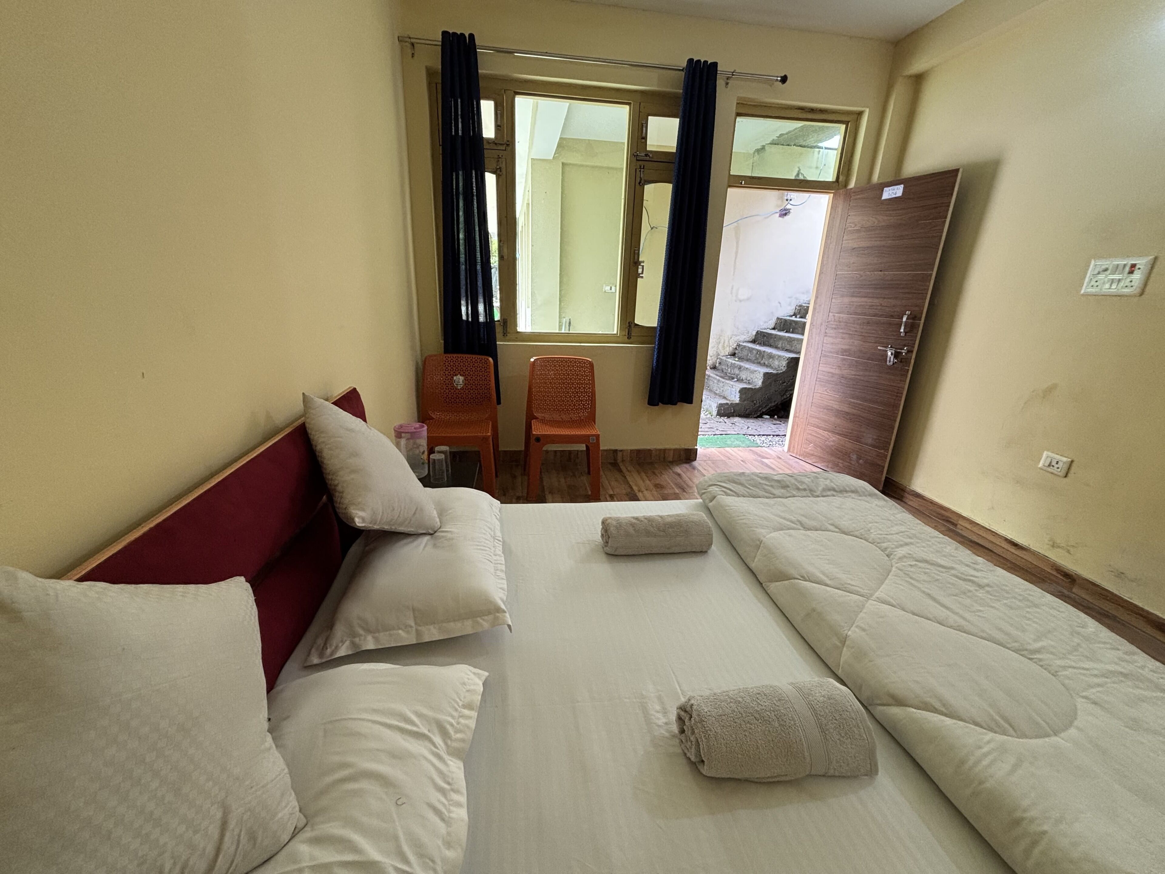 Quarto de hotel com uma cama, duas cadeiras, uma janela com cortinas e uma porta que dá para o exterior.