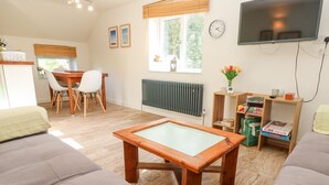 Uma sala de estar aconchegante com mesa de centro de madeira, sofás cinzas, um espaço de jantar com mesa e cadeiras brancas, um radiador, uma TV na parede e uma estante com jogos de tabuleiro.