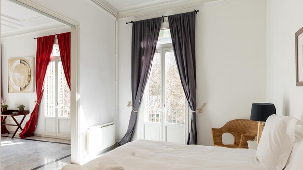 Quarto de hotel com uma cama grande, uma janela com cortinas, um radiador, uma cadeira e um abajur.