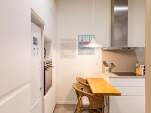 Uma cozinha compacta com armários brancos, forno embutido, coifa de aço inoxidável, uma cadeira de vime, mesa de madeira e luminária fixada na parede.