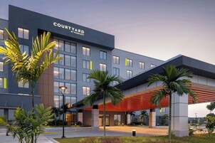 Um hotel Courtyard by Marriott com design moderno, apresentando uma entrada ampla com toldo vermelho, diversas janelas e palmeiras em primeiro plano.