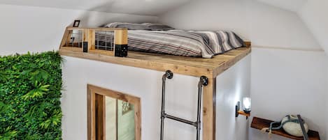 Quarto no estilo loft com cama sobre plataforma de madeira, escada, parede verde e pequena prateleira com luminária e objetos decorativos.