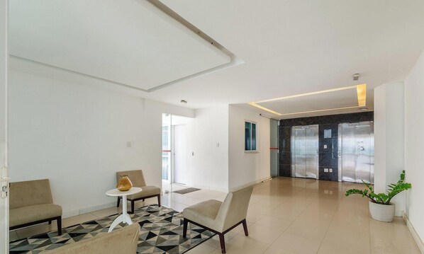 Um hall amplo com design moderno, apresentando um tapete com padrão geométrico, uma área de estar com sofás e uma mesa pequena, e um vaso de planta.