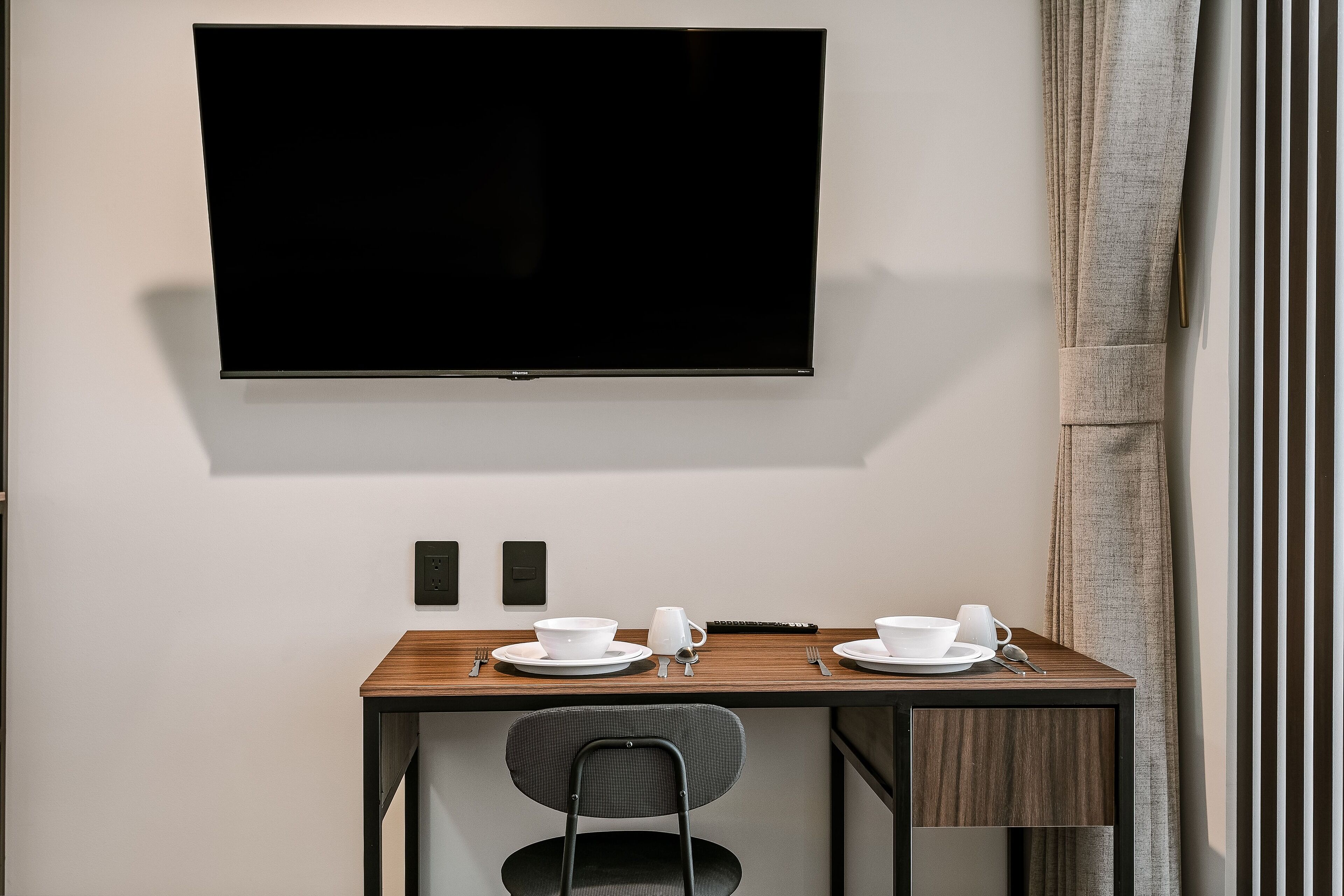 Quarto de hotel com televisão de tela plana na parede, escrivaninha de madeira com cadeira e uma mesa pequena com xícaras e pires brancos.
