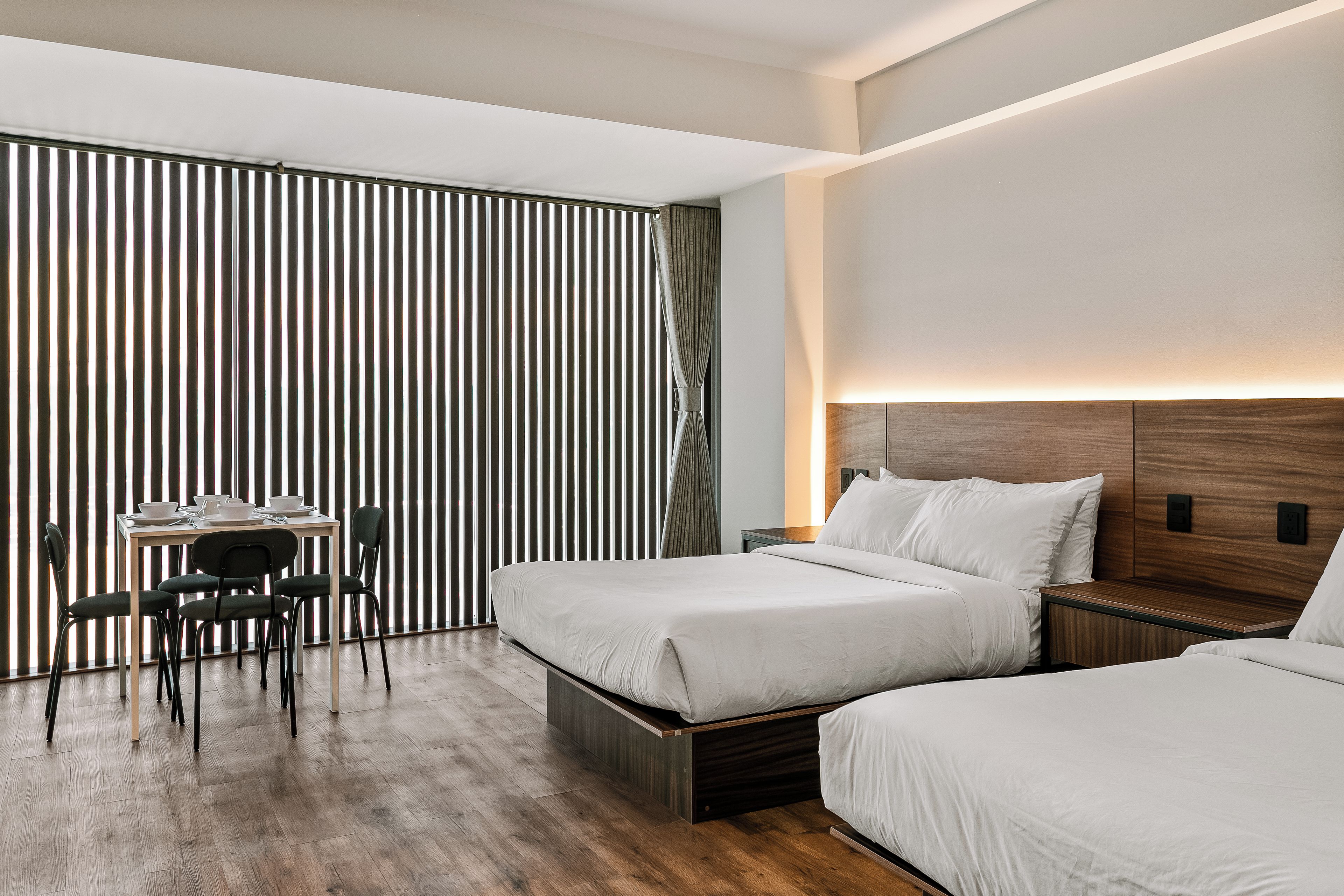 Quarto de hotel com duas camas, uma mesa de jantar com cadeiras e um painel riscado.