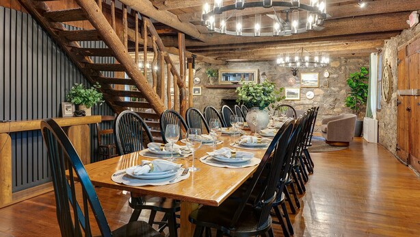 Uma sala de jantar com mesa de madeira posta para seis pessoas, rodeada por cadeiras, um candelabro e uma escadaria.