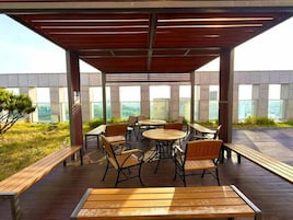 Um pátio externo com mesas e cadeiras de madeira, um banco de madeira e vista para o horizonte urbano através de grandes janelas.