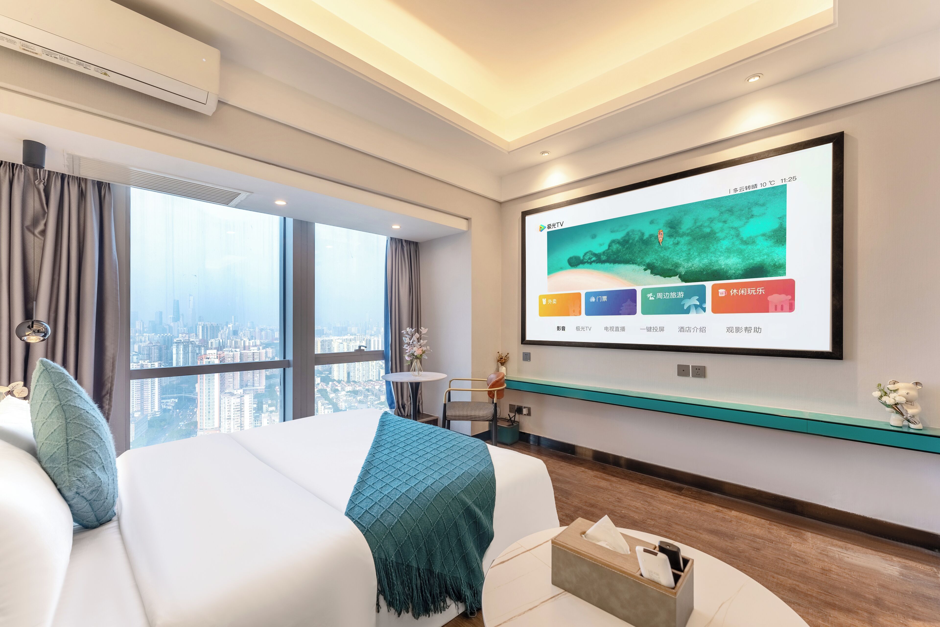 Quarto de hotel com uma cama grande, televisão fixada na parede, vista da cidade pela janela e uma mesinha com um vaso de flores.