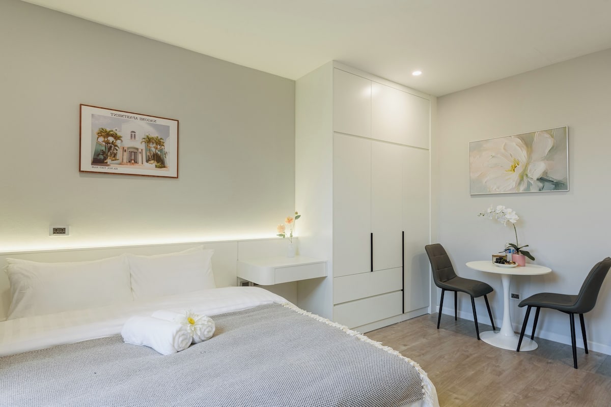 Quarto de hotel moderno com cama bem arrumada, uma pequena escrivaninha com um vaso de flores, uma mesa redonda com duas cadeiras e um guarda-roupa grande.