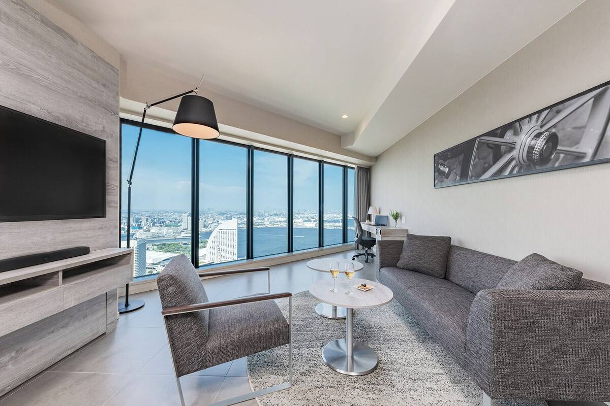 Uma sala de estar moderna com uma televisão grande, assentos confortáveis, uma mesa redonda pequena e vista para a cidade.