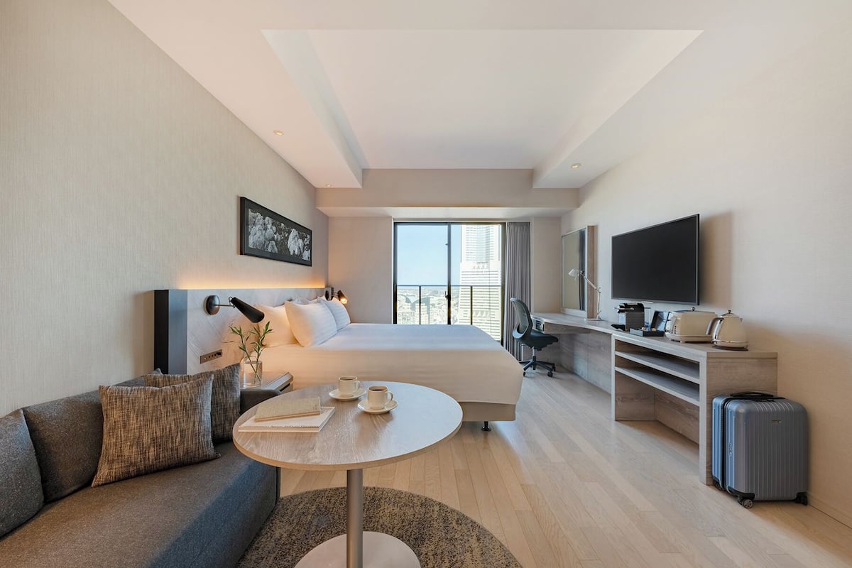 Quarto de hotel moderno com cama, sofá, escrivaninha com televisão, mesa de centro e mala.