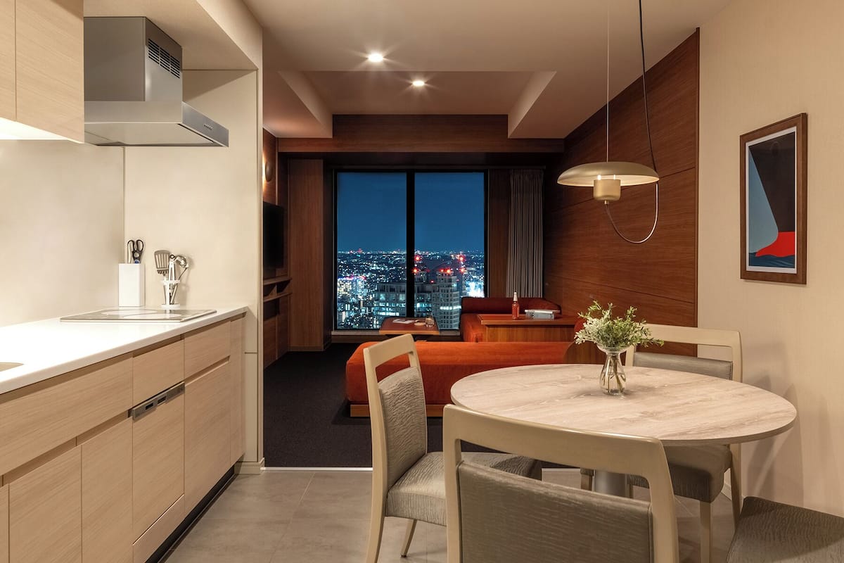 Quarto de hotel moderno com cozinha, área de refeições e uma grande janela com vista para a cidade.