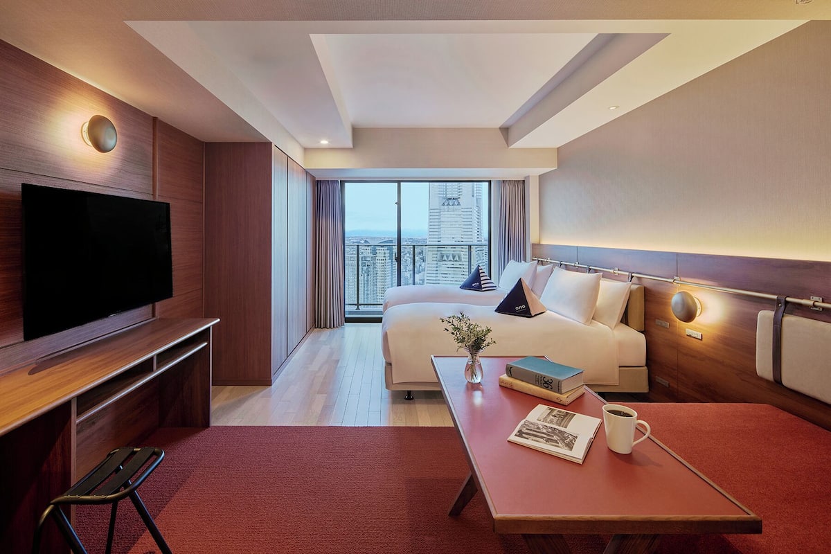 Quarto de hotel com televisão de tela plana grande, uma cama com almofadas, mesa de cabeceira com um livro e uma caneca, e vista para a cidade através de grandes janelas.