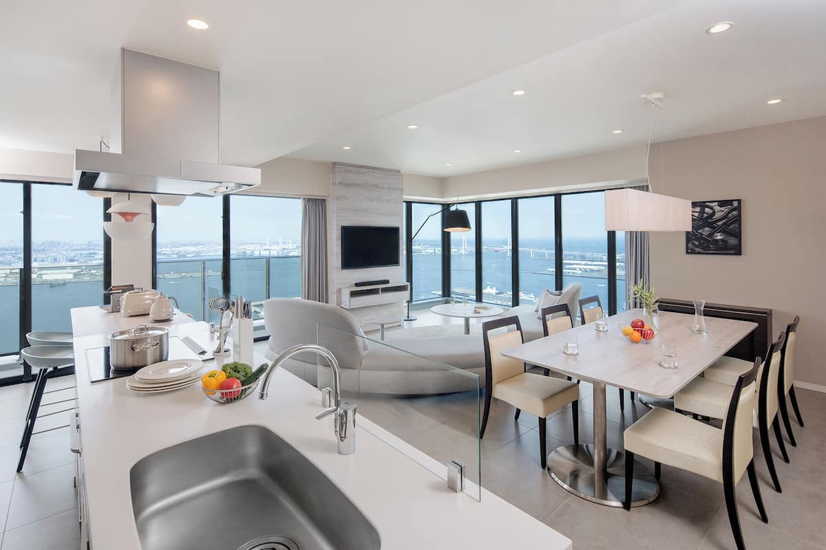 Cozinha moderna com uma ilha grande, área de jantar com mesa redonda e sala de estar com televisão e grandes janelas com vista para a paisagem urbana.